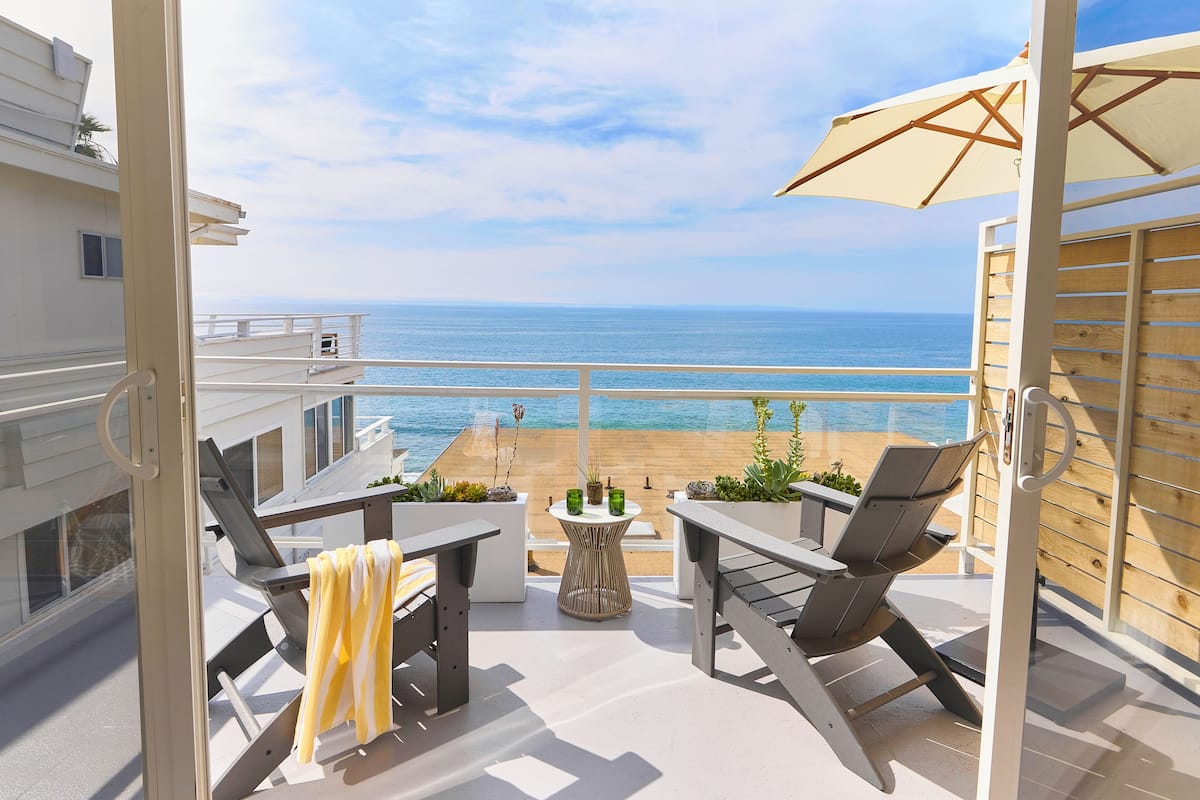 Uma varanda com vista para o mar, equipada com cadeiras, uma mesa e um guarda-sol.