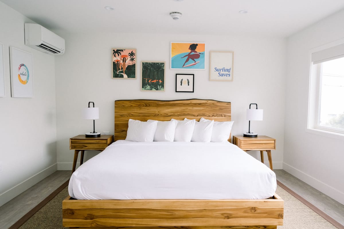 Quarto com cama de madeira, roupa de cama branca, duas mesas de cabeceira com luminárias e obras de arte na parede, incluindo um cartaz com a inscrição 'Surfing Saves'.