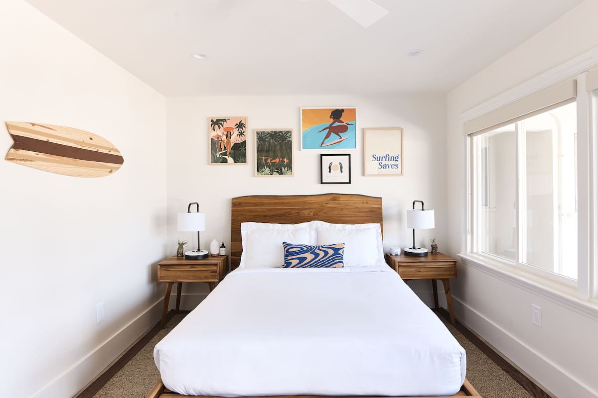 Quarto bem arrumado com uma cama grande, duas mesas de cabeceira com luminárias e obras de arte nas paredes retratando surf e paisagens tropicais.