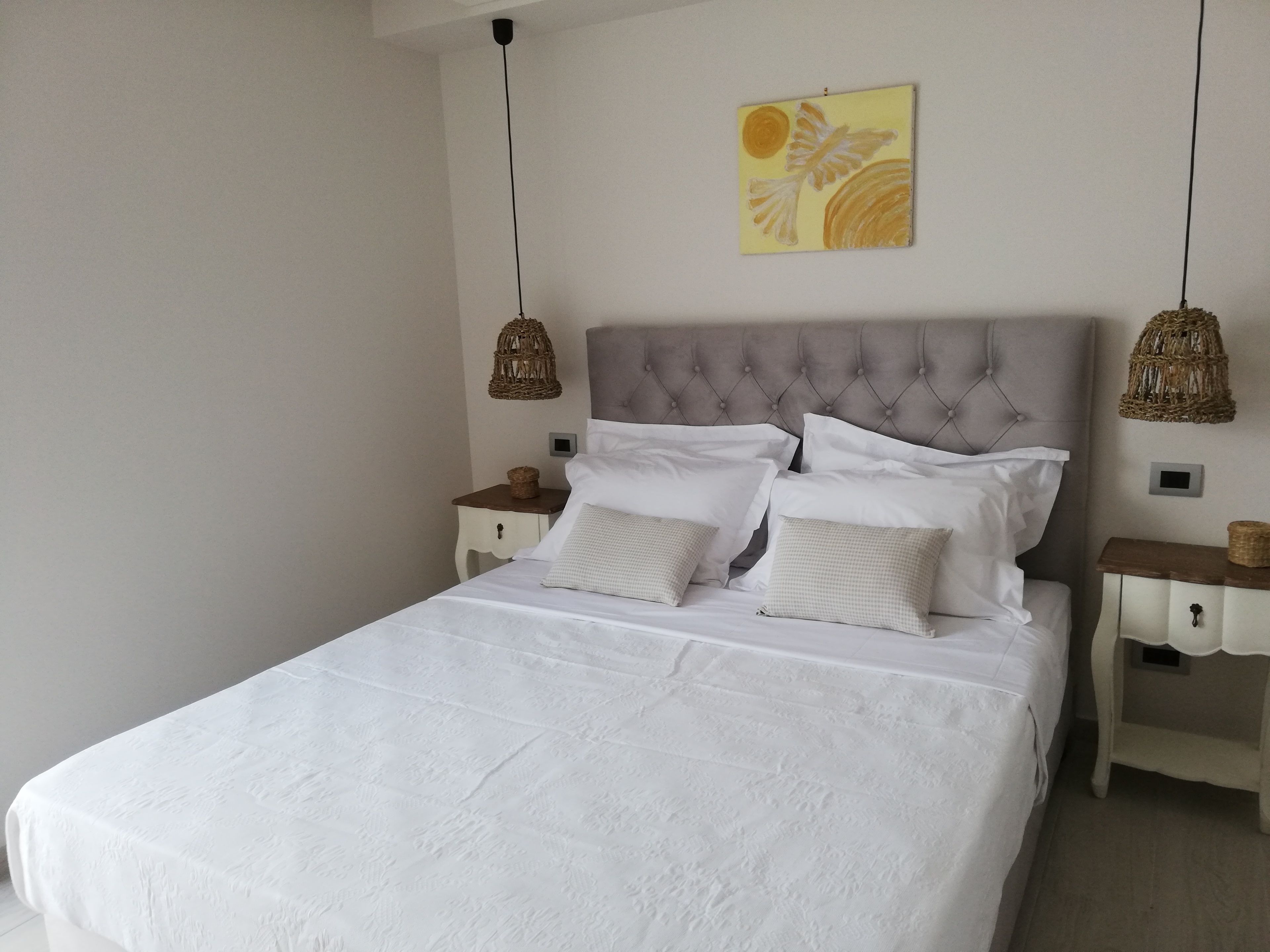 Cama bem arrumada com lençóis brancos, cabeceira estofada e duas mesas de cabeceira com abajures de vime.
