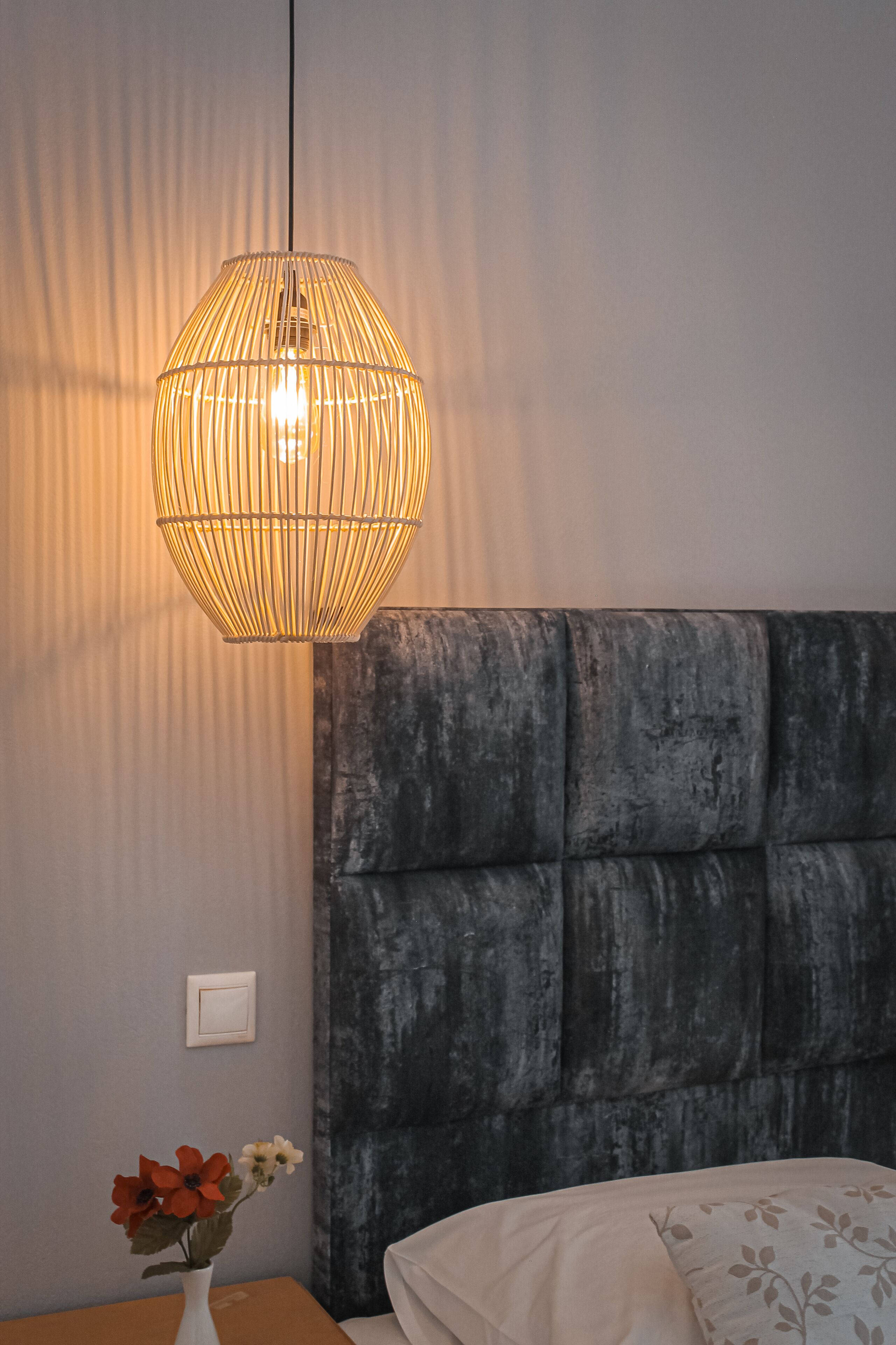 Quarto com cabeceira cinza texturizada, mesa de cabeceira com um vaso de flores e luminária pendente.