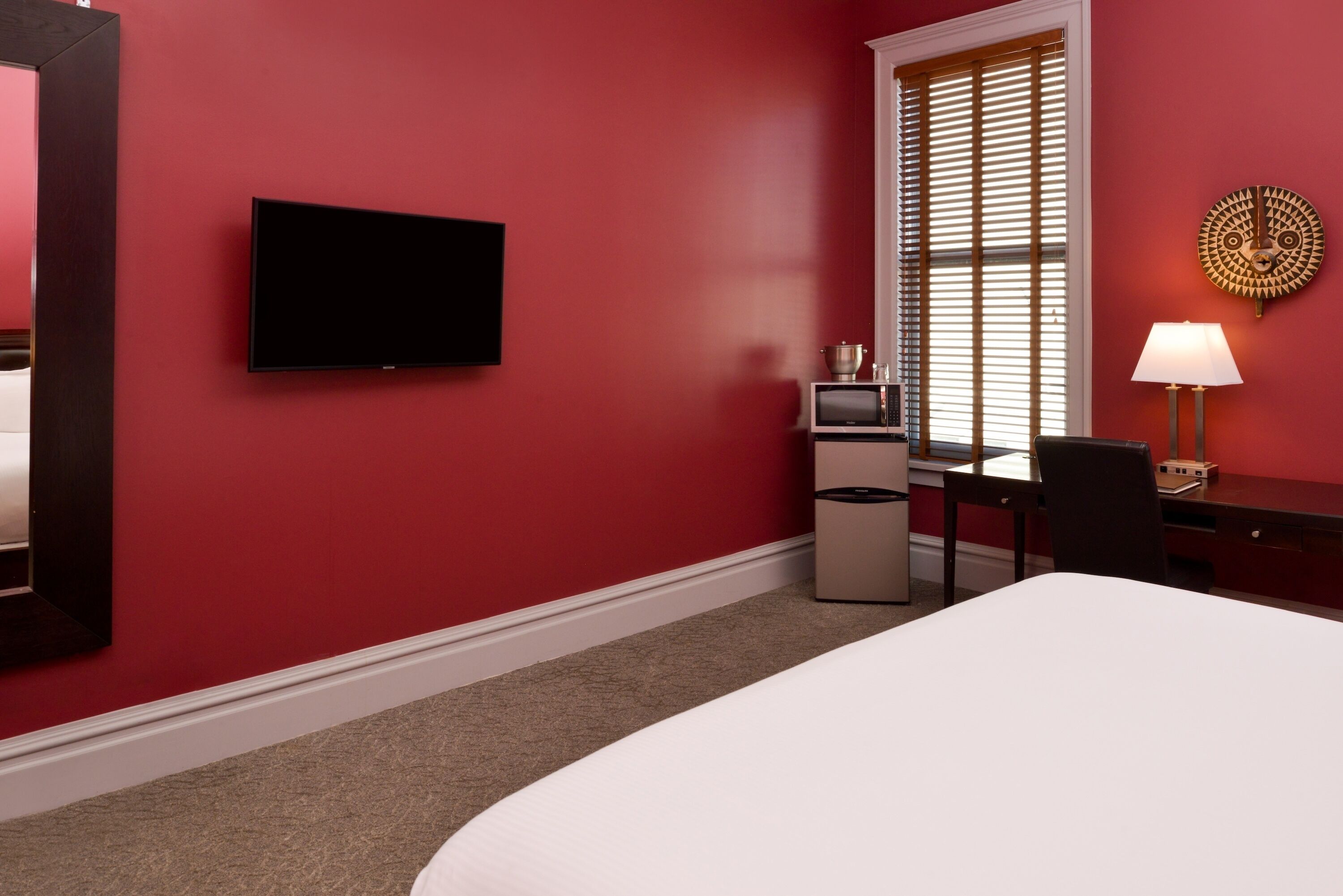 Quarto de hotel com uma cama grande, televisão na parede, secretária com abajur e janela com persianas.