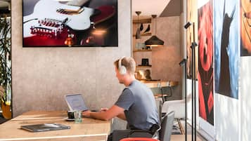 Uma pessoa trabalhando em uma mesa com um laptop, fones de ouvido e um livro. Uma guitarra está exposta na parede, e há cartazes e prateleiras com diversos objetos.