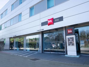 Um edifício moderno com fachada de vidro extensa, um grande letreiro vermelho da marca 'ibis' e vista clara para o estacionamento.