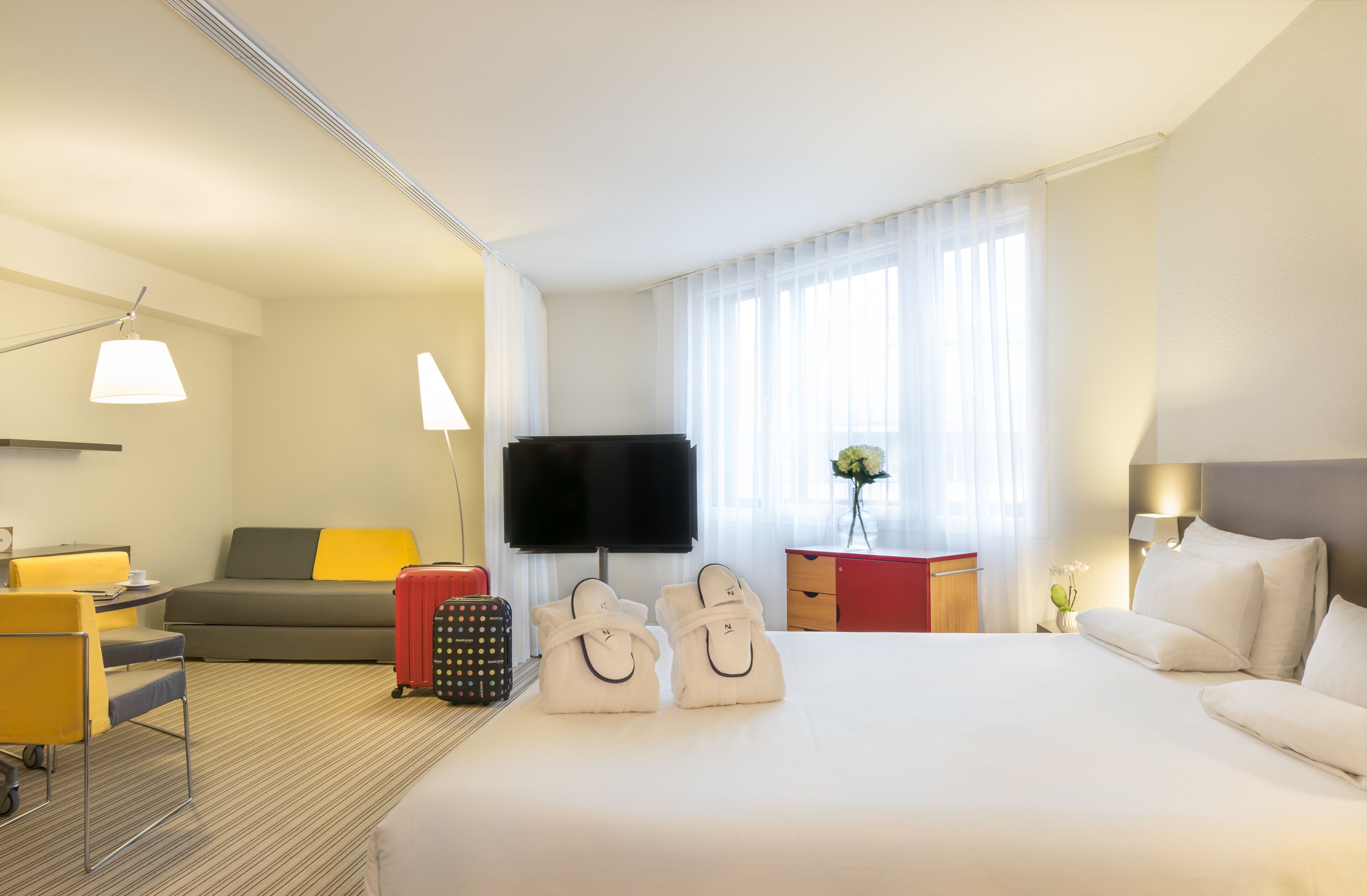 Quarto de hotel com cama, escrivaninha, cadeira, televisão e janela com cortinas.