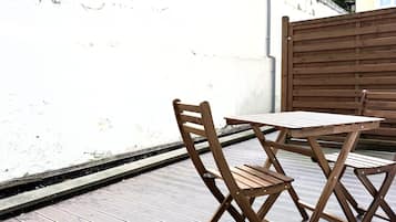Mesa e cadeiras de madeira em um deck de madeira, com uma parede branca e uma cerca de madeira ao fundo.