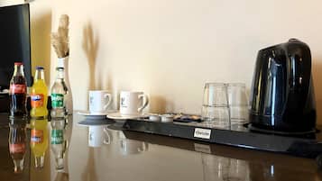 Espaço de refeições em quarto de hotel, com cafeteira, xícaras e garrafas de bebidas.