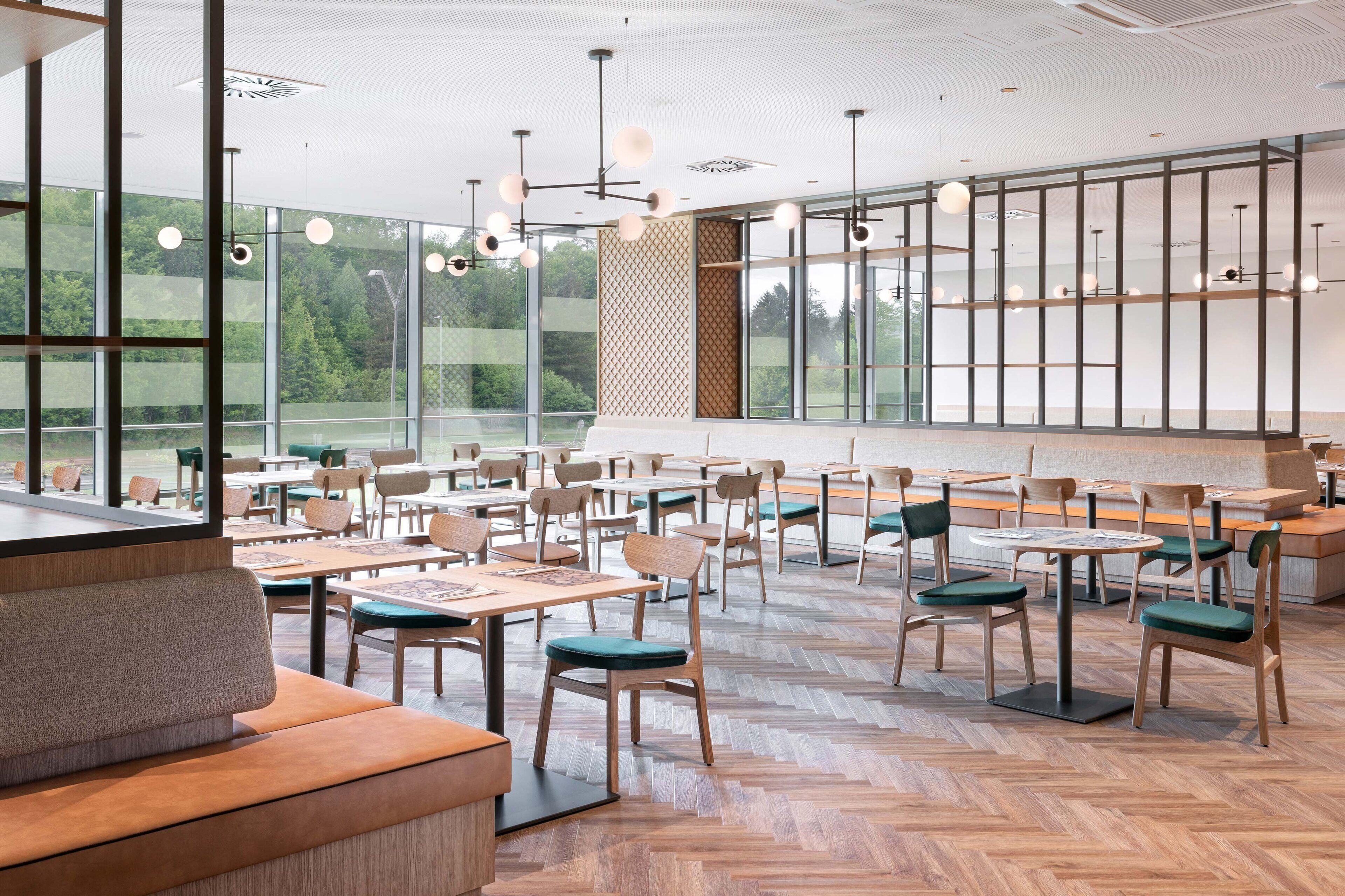 Um espaço amplo e interno com várias mesas e cadeiras, grandes janelas com vista para o verde lá fora e um design de teto moderno com luminárias pendentes.
