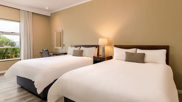 Quarto de hotel com duas camas, uma escrivaninha, uma cadeira, uma mesa de cabeceira, uma luminária, um espelho e uma janela com vista.