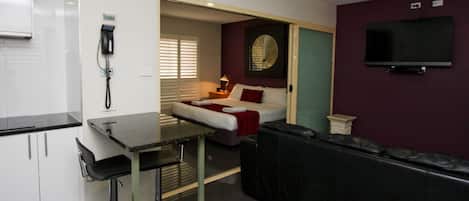 Quarto de hotel compacto com cozinha compacta, uma cama com roupa de cama vermelha, televisão presa à parede e uma pequena área de estar.