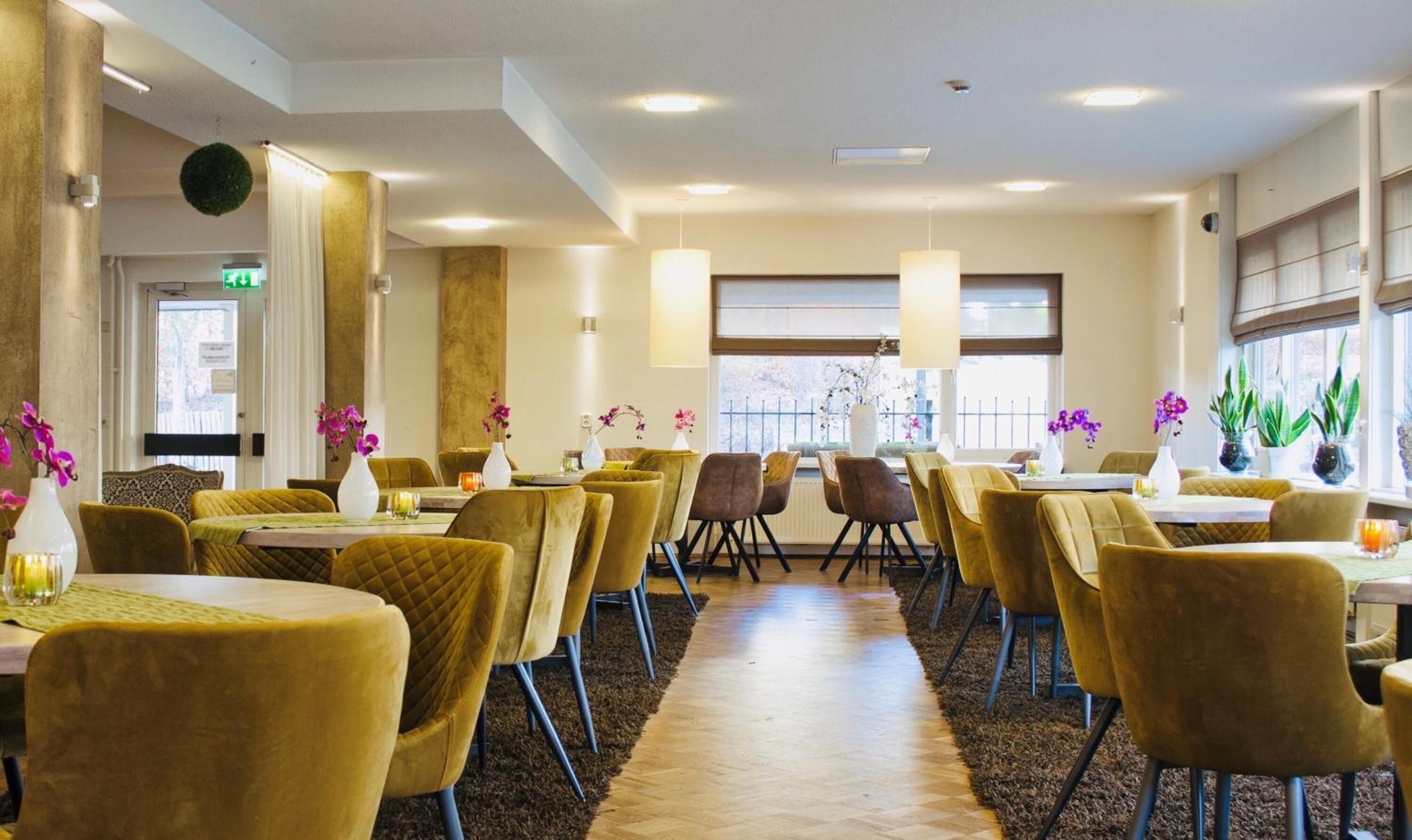 Um restaurante com várias mesas e cadeiras, piso de madeira e janelas amplas.