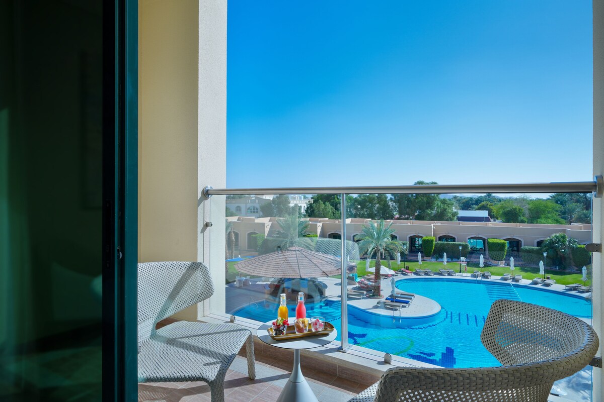Varanda com cadeiras de vime, uma mesa pequena com bebidas e vista para a piscina e área do resort.