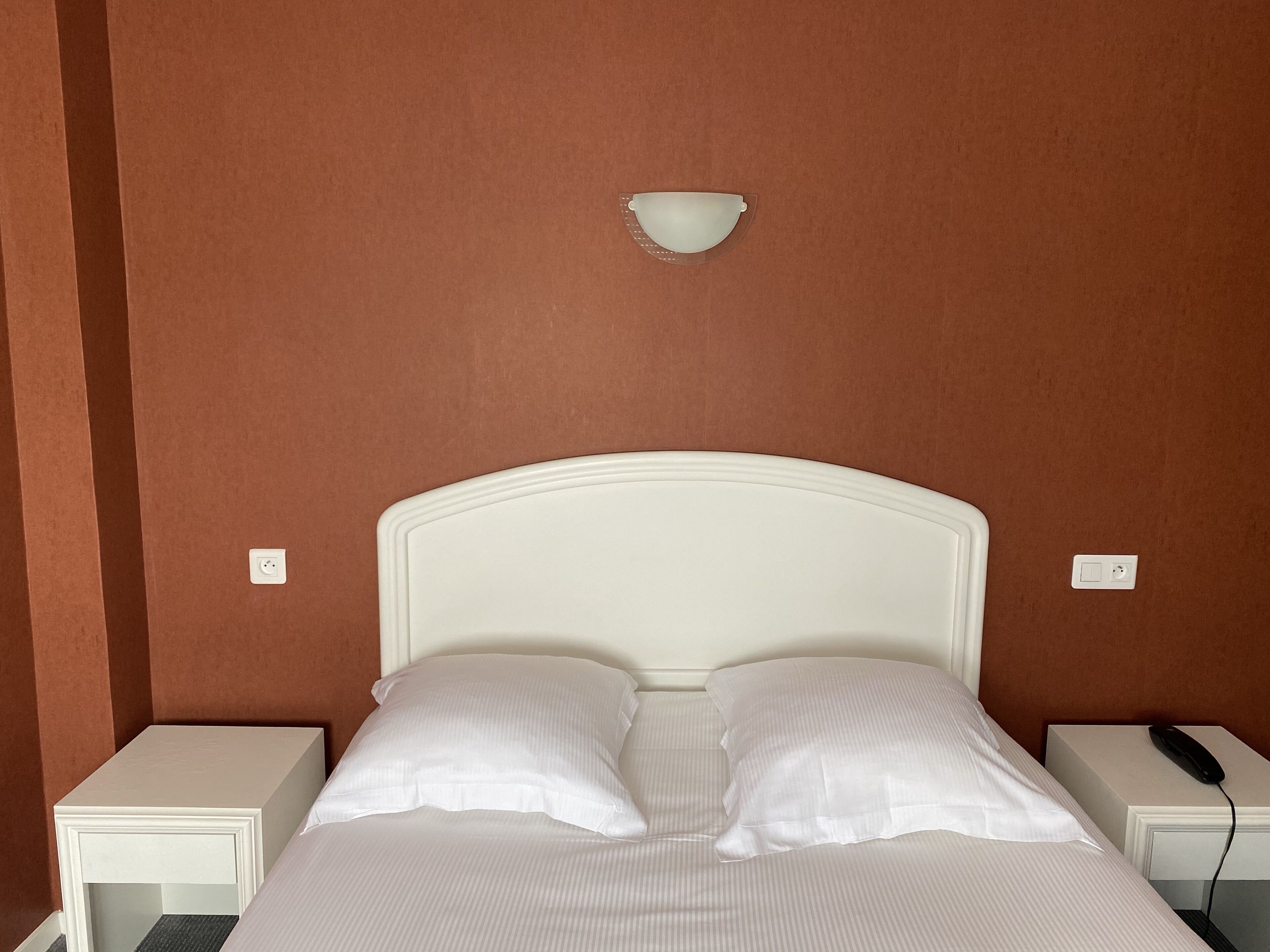 Cama bem arrumada com lençóis e almofadas brancas, duas mesas de cabeceira com abajures e uma tomada na parede.
