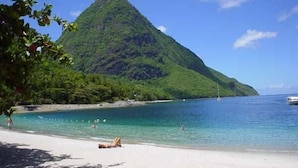 Uma praia com águas turquesa cristalinas, uma pessoa bronzeando-se e uma montanha ao fundo.