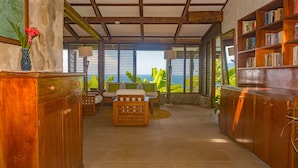 Um quarto com teto de madeira, uma estante de livros, uma sala de jantar com mesa e cadeiras, e vista para o oceano.