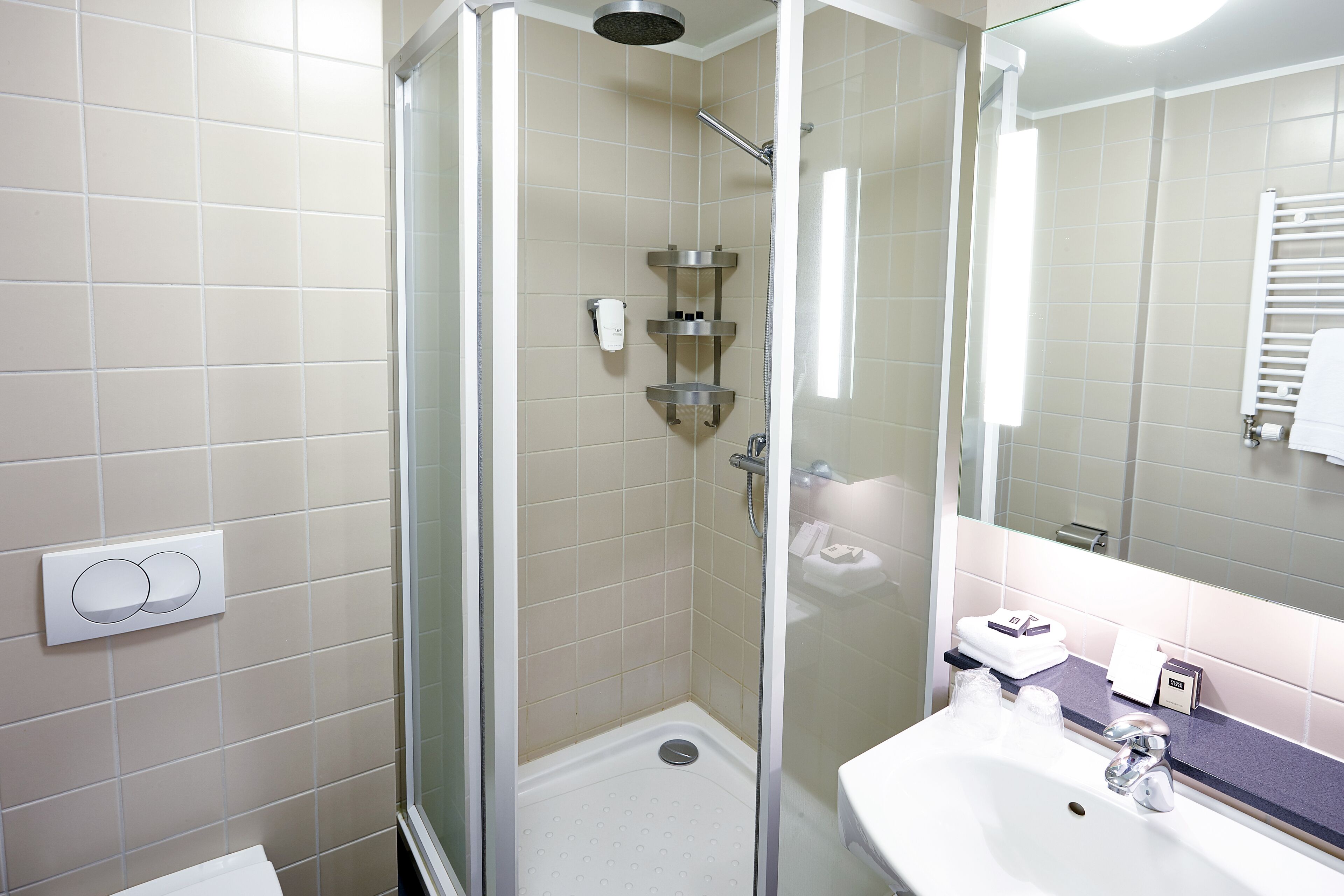 Casa de banho moderna com chuveiro separado por vidro, lavatório branco e espelho a refletir um porta-toalhas.