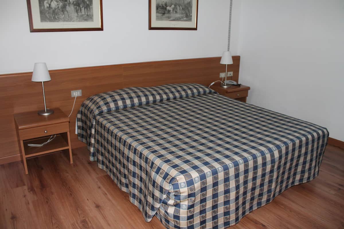 Quarto com cabeceira de madeira, colcha xadrez, mesa de cabeceira com abajur e criado-mudo com telefone.