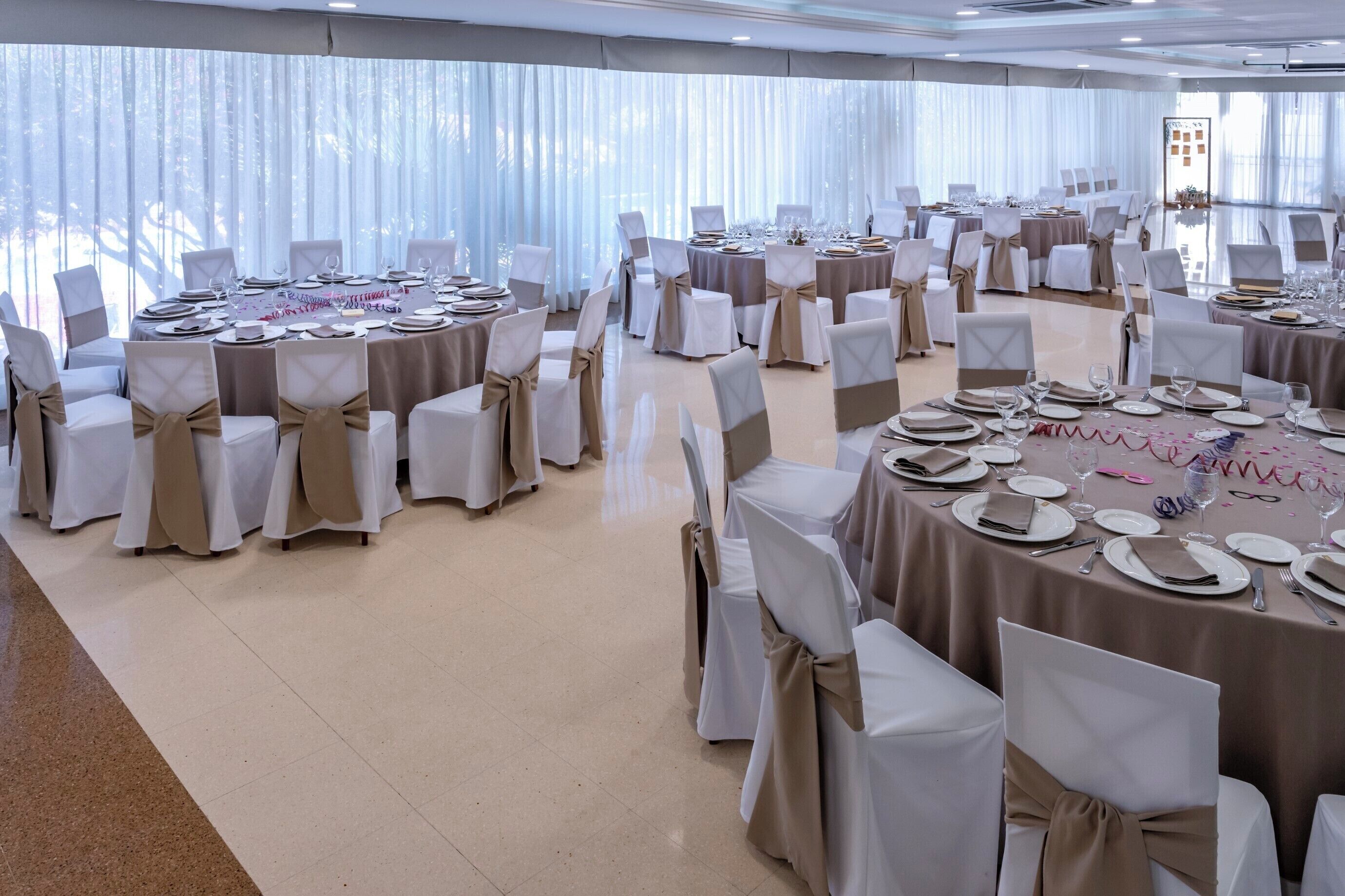 Uma sala de banquetes com mesas redondas preparadas para refeições, cadeiras brancas e bege, e uma grande janela com cortinas leves.