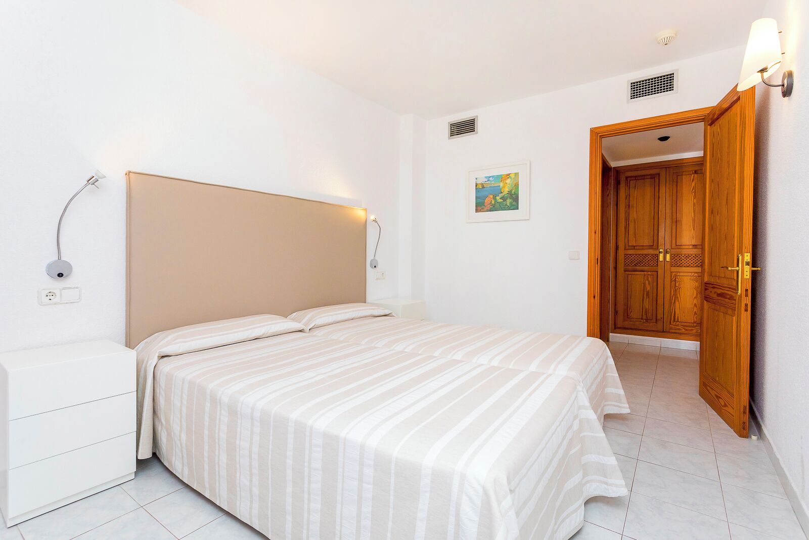 Quarto com cama, mesa de cabeceira, luminária na parede e porta para outro cômodo.