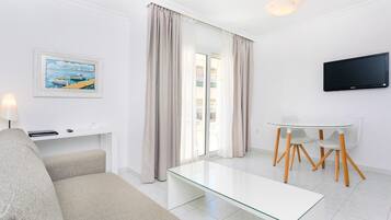 Uma sala de estar moderna com uma mesa de centro de vidro, um sofá, uma TV na parede e um quadro de uma paisagem costeira.