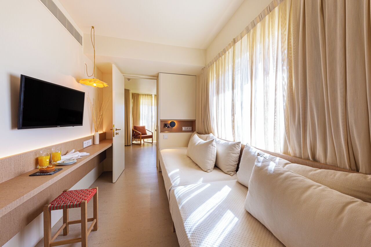 Quarto de hotel com sofá, televisão na parede, uma mesa pequena com cadeira e janela com cortinas.