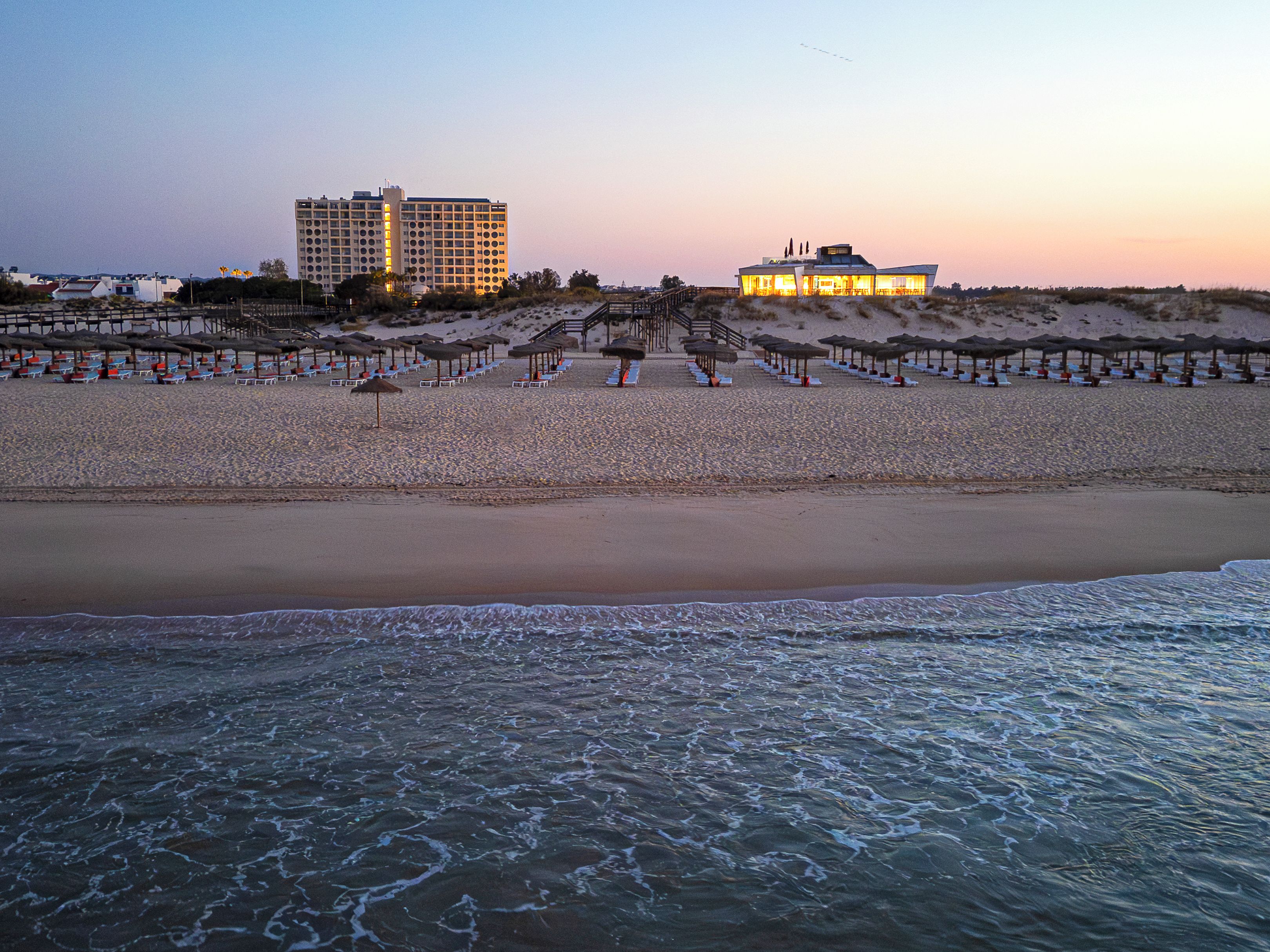 Uma praia com fileiras de guarda-sóis e espreguiçadeiras, um prédio com varanda voltada para a praia e uma área de piscina de hotel.