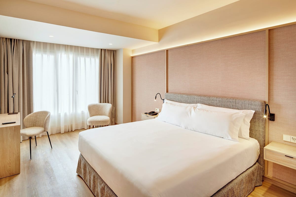 Quarto de hotel com uma cama grande, uma escrivaninha com cadeira, uma televisão e uma janela com cortinas.