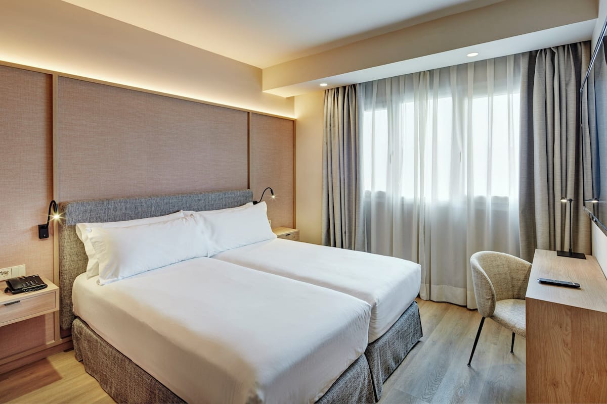 Quarto de hotel com cama de casal, mesas de cabeceira, escrivaninha com cadeira, televisão e janela com cortinas.