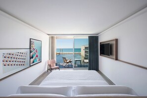 Quarto de hotel com uma cama grande, televisão, quadro emoldurado e vista para o mar através de uma porta deslizante.
