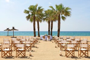 Decoração de casamento na praia, com fileiras de cadeiras de madeira e uma passagem central até um palco, palmeiras e o oceano ao fundo.