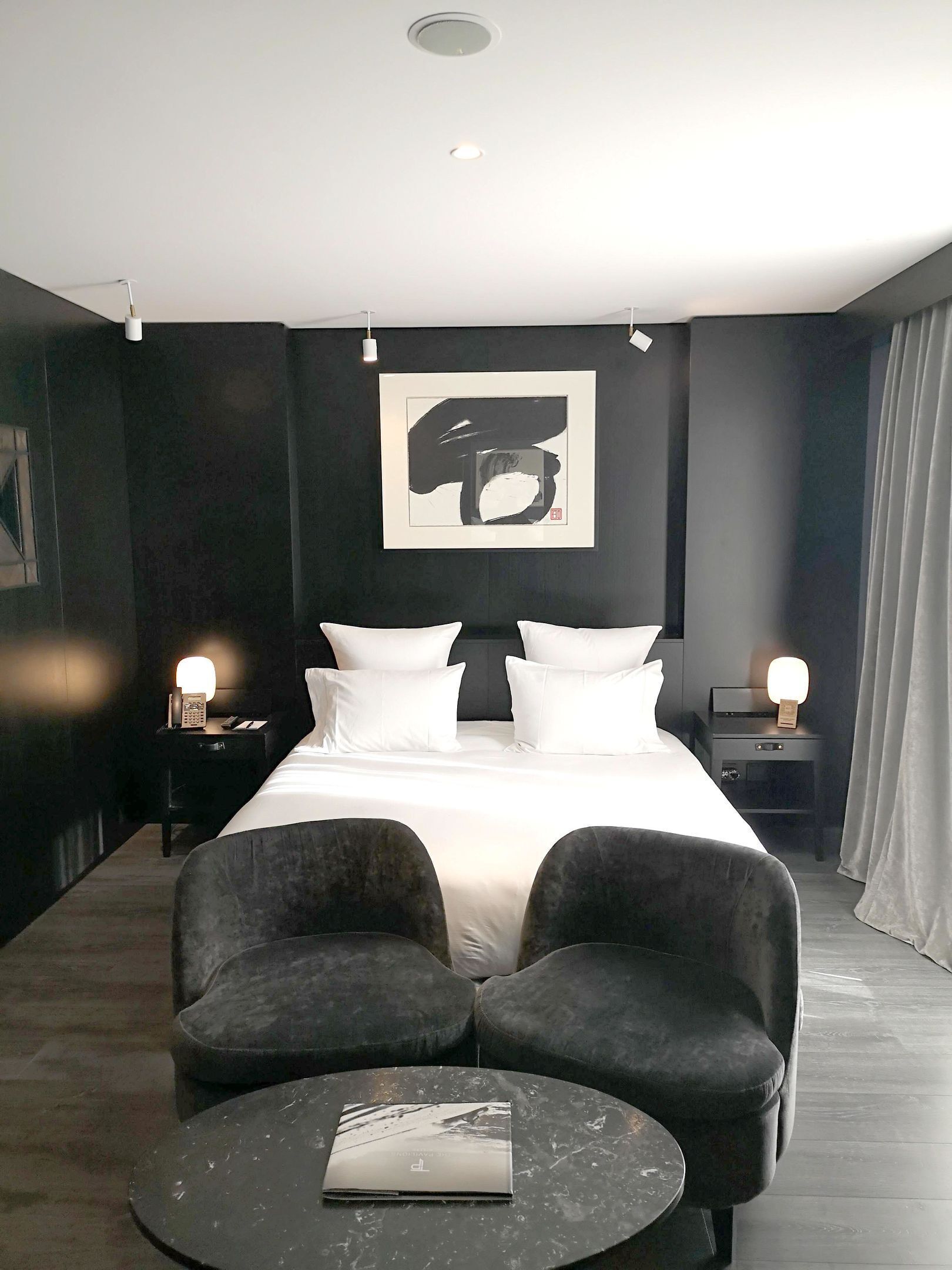 Quarto de hotel moderno com uma cama grande, duas poltronas, uma mesa redonda pequena e um quadro na parede.