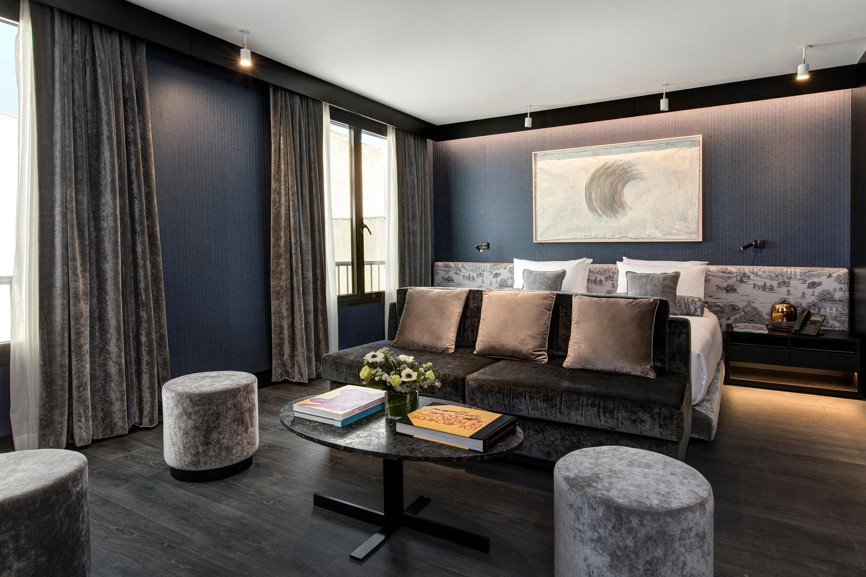 Uma sala de estar moderna com uma parede azul escura de destaque, um grande quadro abstrato, um sofá com diversas almofadas, uma mesa de centro redonda com livros e um vaso de flores, e dois pufes redondos.