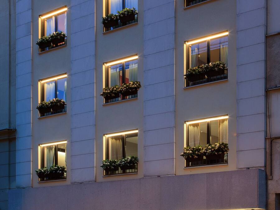 Um hotel moderno com vários andares, cada um com varandas adornadas com plantas em vasos. O edifício tem um design contemporâneo e elegante, com grandes janelas e fachada bem iluminada.