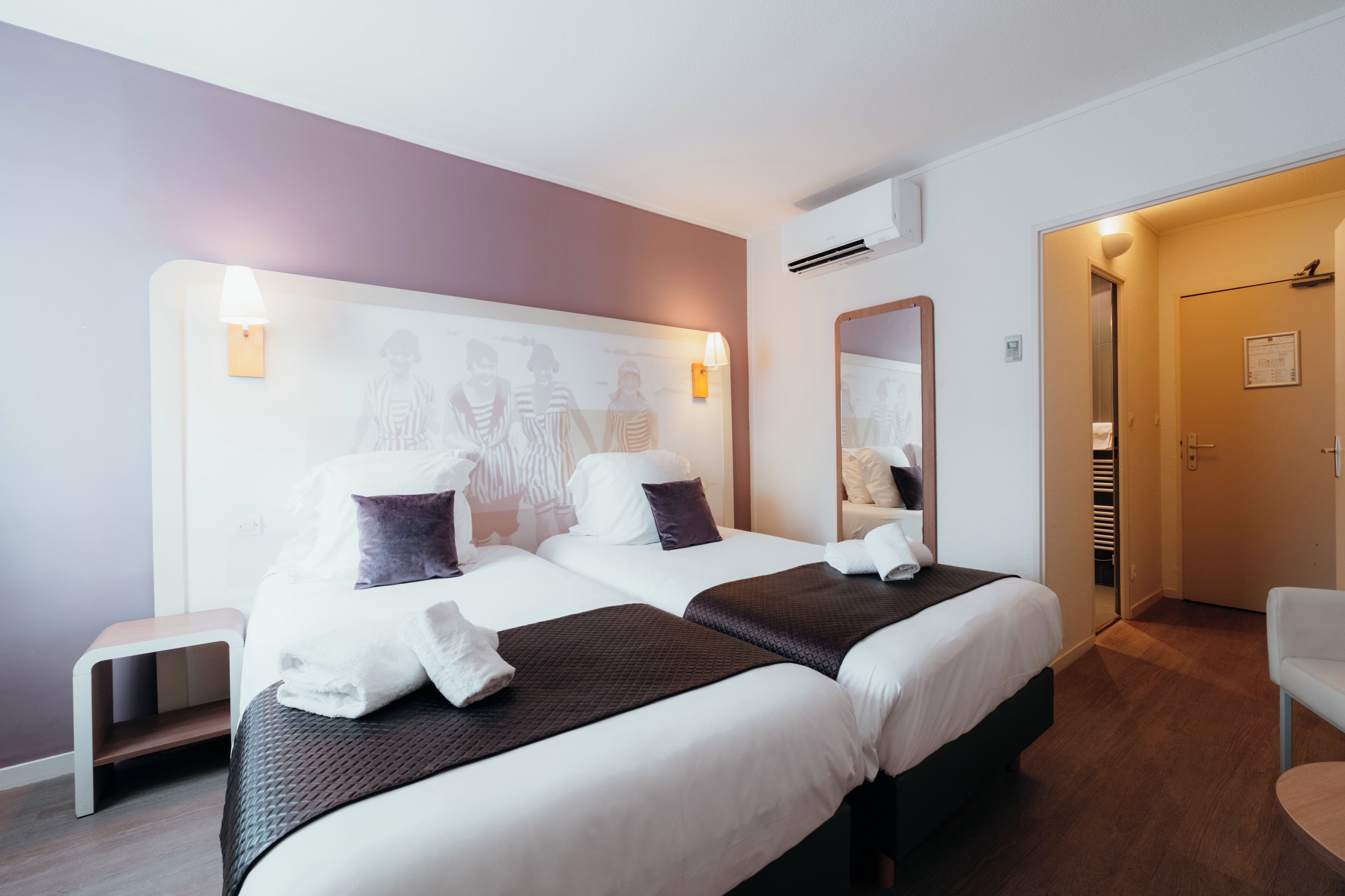 Quarto de hotel com duas camas, uma mesinha, uma cadeira, um espelho e um aparelho de ar condicionado na parede.