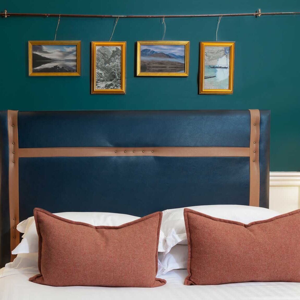 Uma cama com cabeceira azul escura, roupa de cama branca e duas almofadas castanhas. Quatro quadros emoldurados estão pendurados na parede acima da cama.