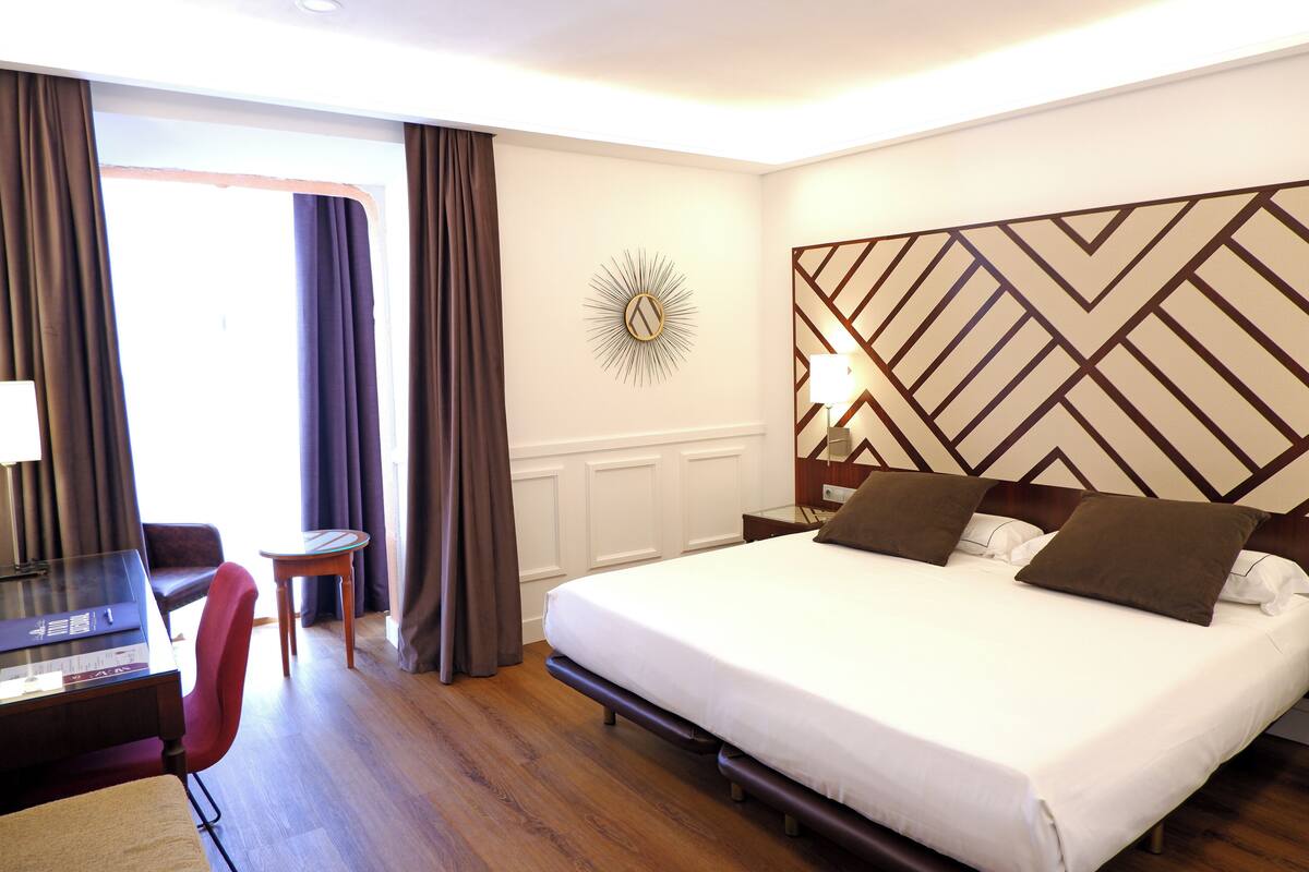 Quarto de hotel com uma cama grande, uma escrivaninha com cadeira, uma mesinha e uma janela com cortinas.