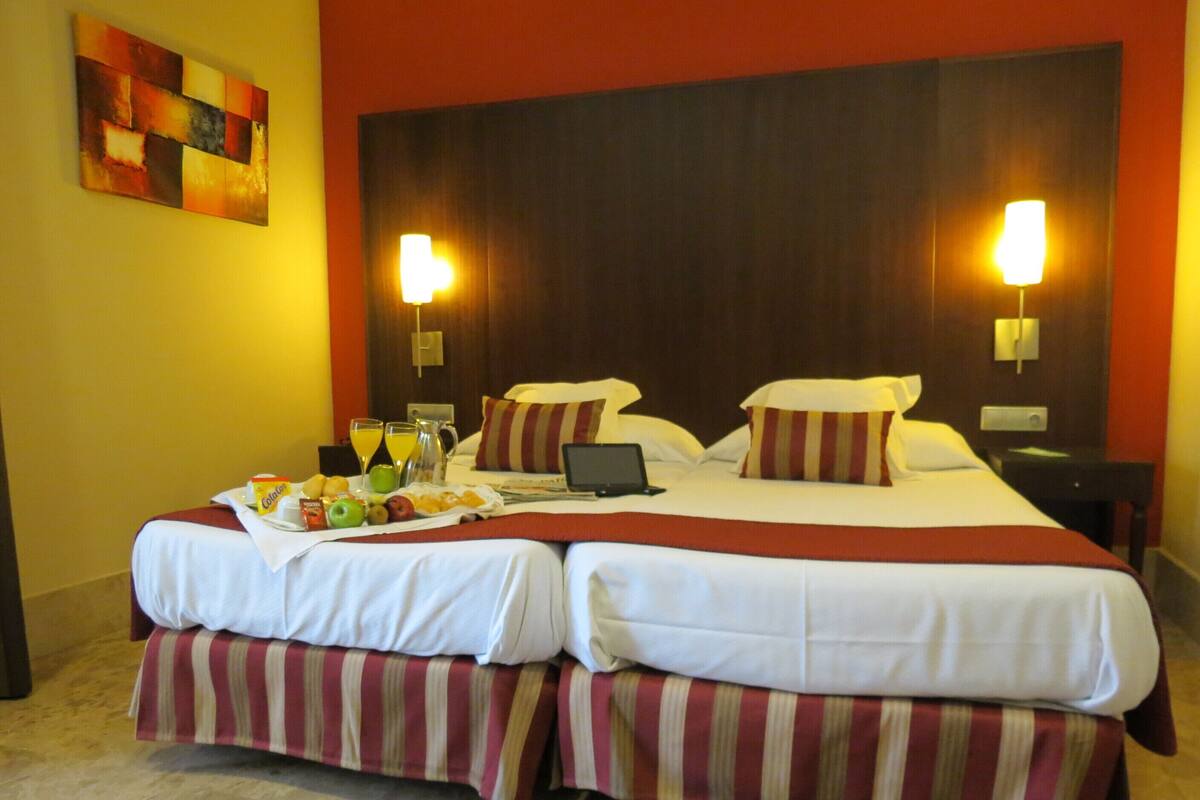 Uma cama de casal com saia de cama listrada em vermelho e branco, um laptop na cama e uma bandeja de café da manhã com frutas e suco.