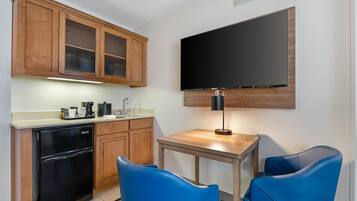 Cozinha compacta com geladeira, cafeteira e armários. Há uma TV de tela plana na parede e uma mesa pequena com duas cadeiras.