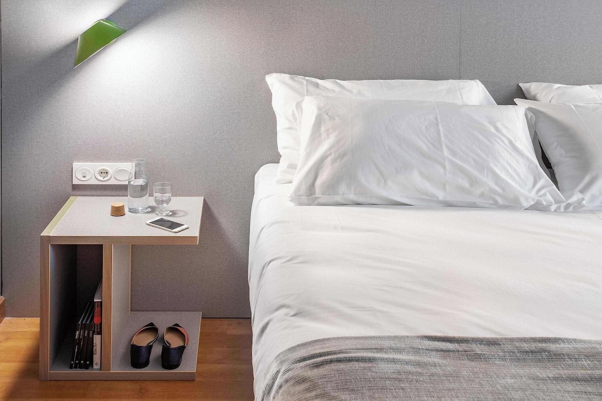Cama bem arrumada com roupa de cama branca, mesa de cabeceira com abajur, um copo de água, um smartphone e um par de sapatos no chão.