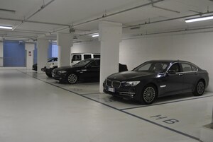 Uma garagem subterrânea com paredes brancas e piso de concreto, com vários carros estacionados, incluindo um BMW preto.