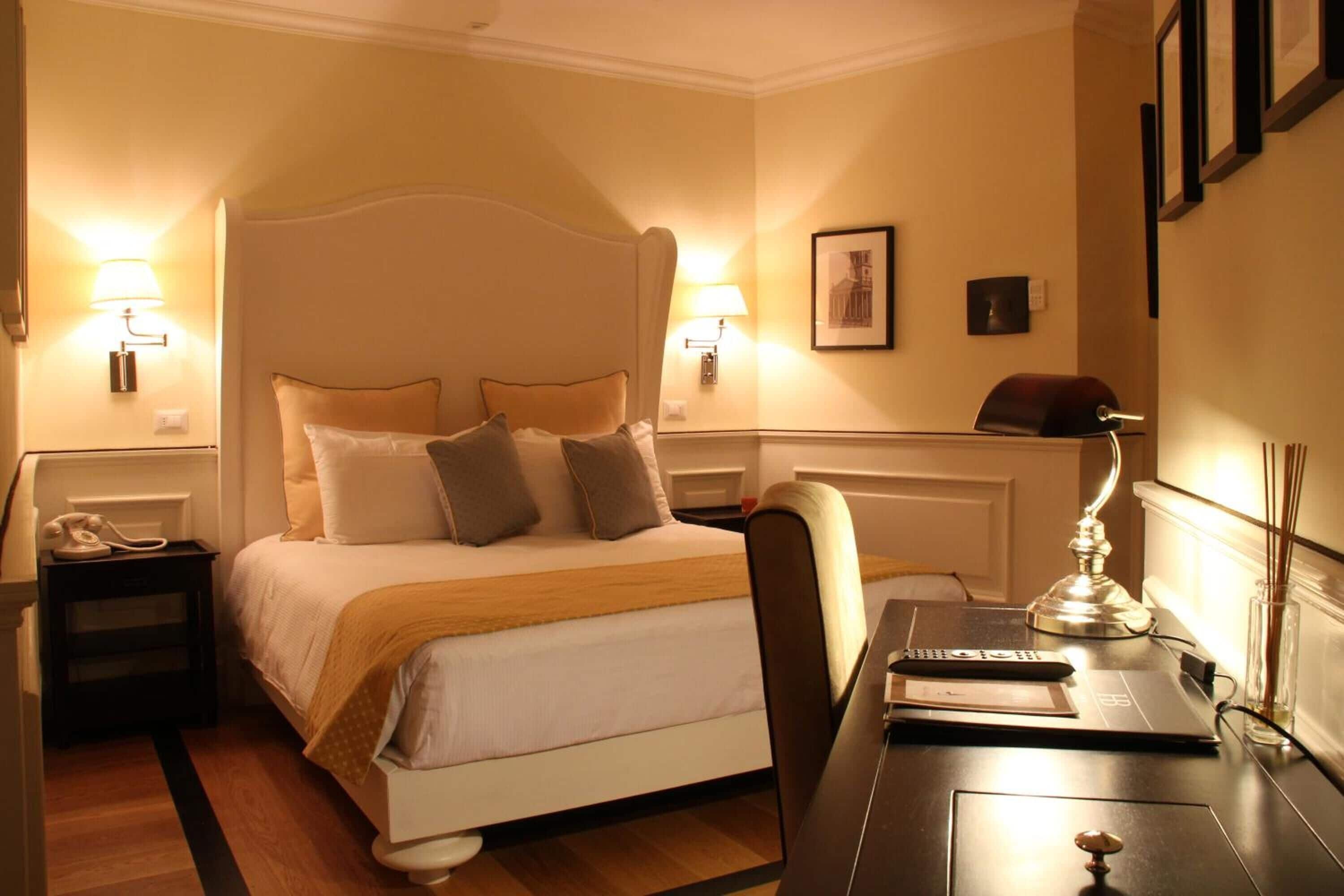 Quarto de hotel com uma cama grande, abajures, uma escrivaninha com luminária, uma cadeira e um telefone.