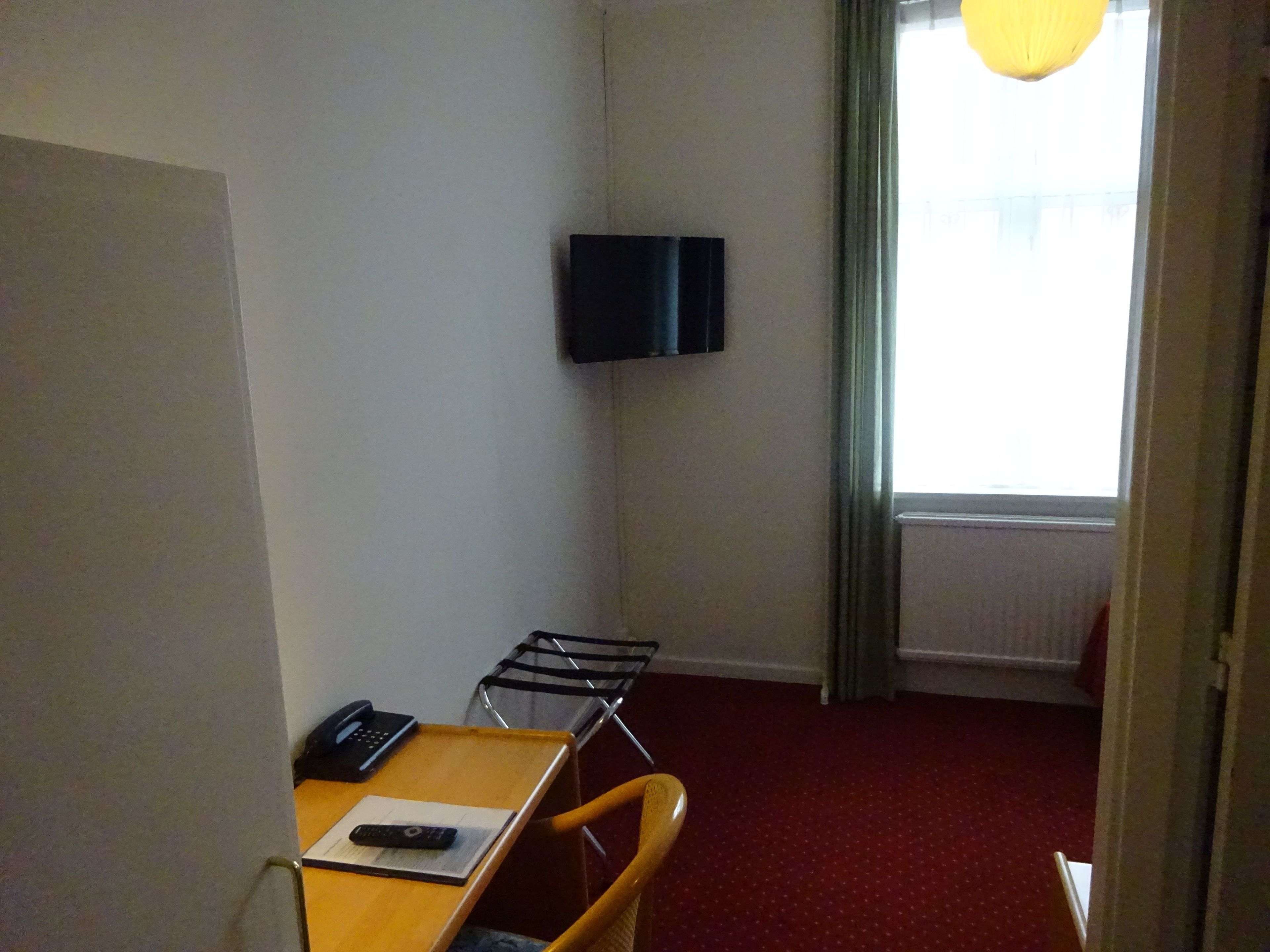 Quarto de hotel com uma cama de solteiro, uma mesinha com uma cadeira, televisão fixada na parede e uma janela com cortinas.