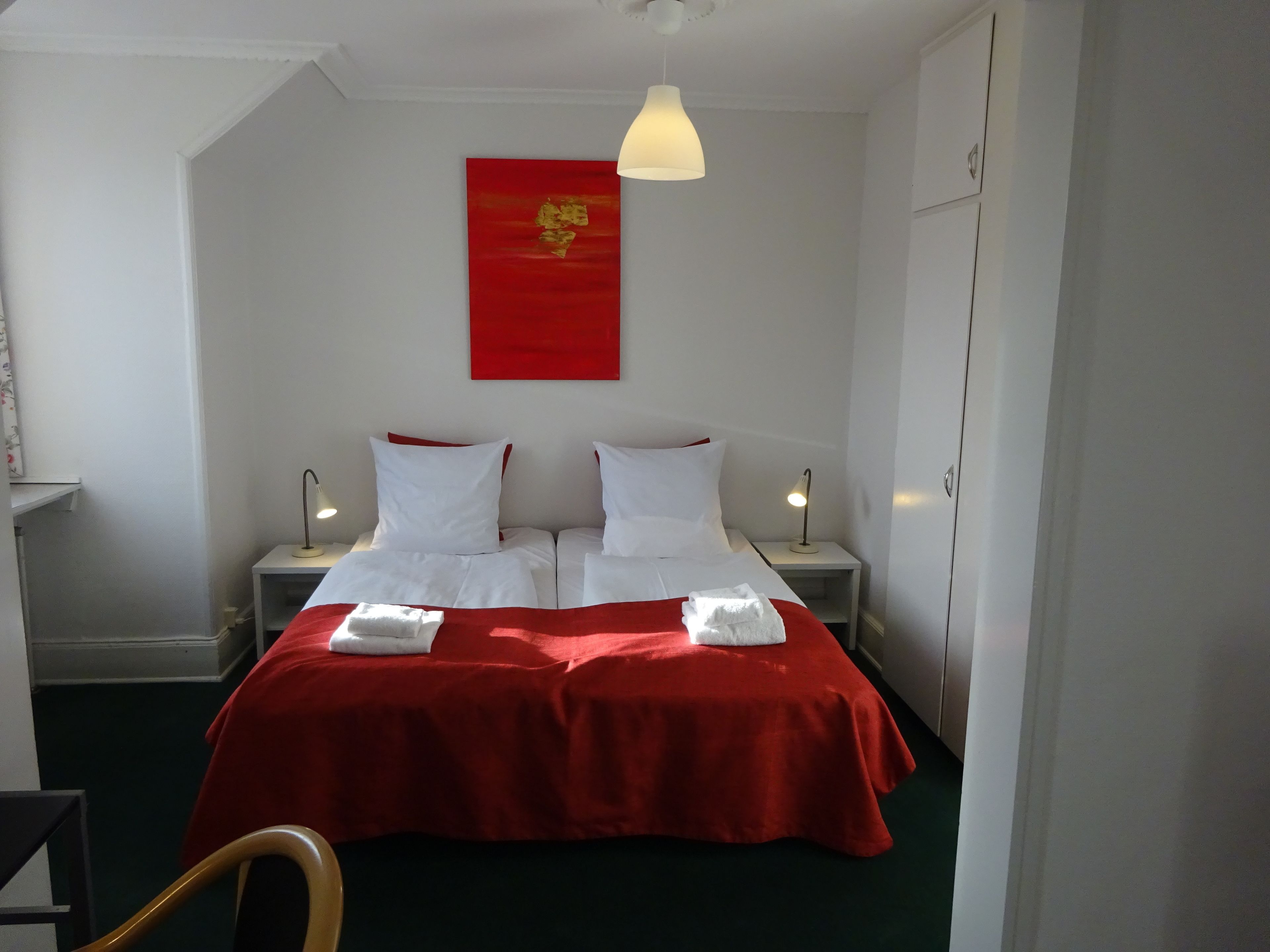 Cama bem arrumada com roupa de cama branca e vermelha, duas luminárias de cabeceira e um tapete de parede vermelho.