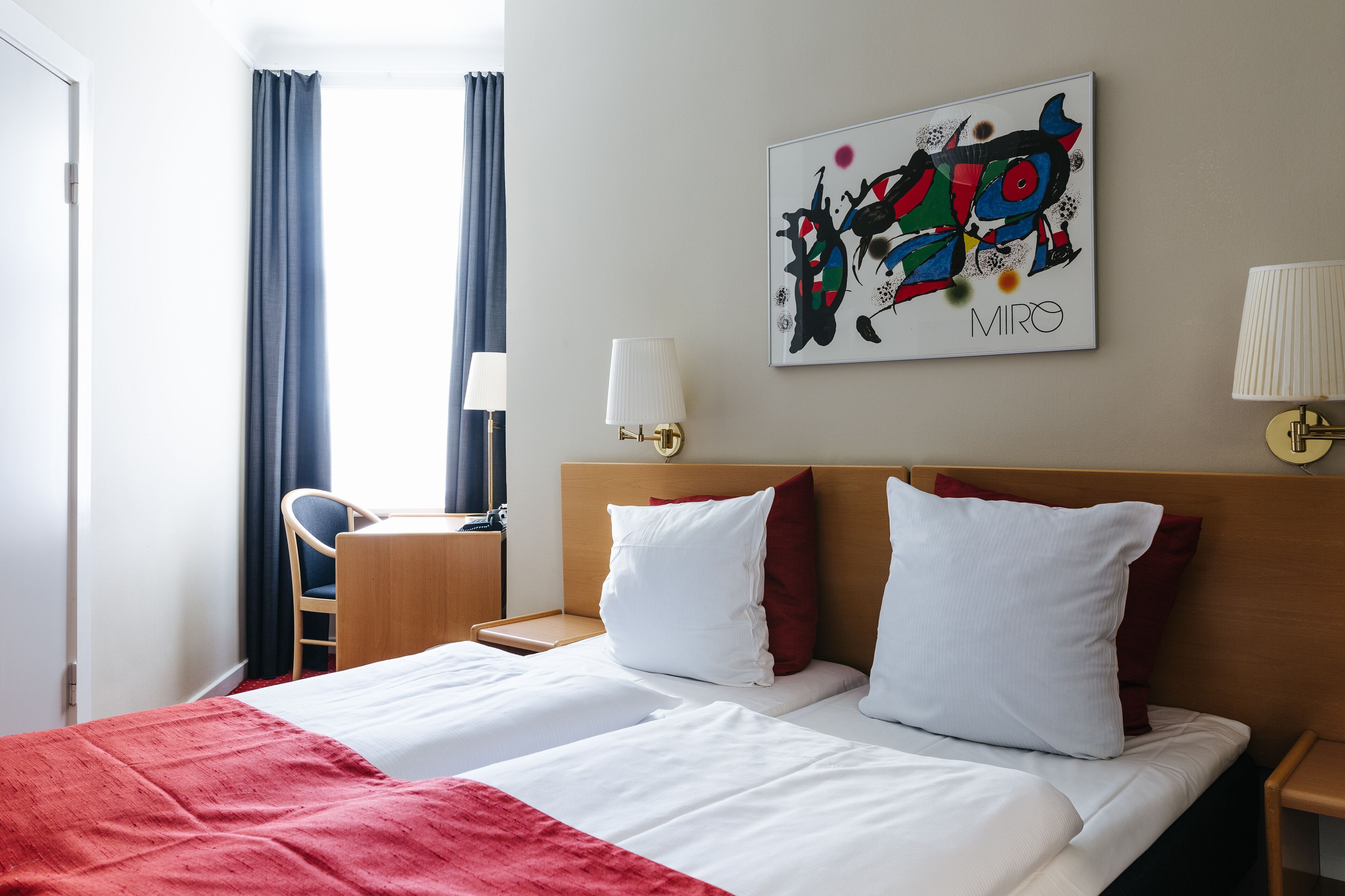 Quarto de hotel com uma cama grande, uma escrivaninha e uma cadeira. Há um quadro na parede e uma janela com cortinas.