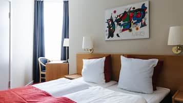Quarto de hotel com uma cama grande, uma escrivaninha e uma cadeira. Há um quadro na parede e uma janela com cortinas.