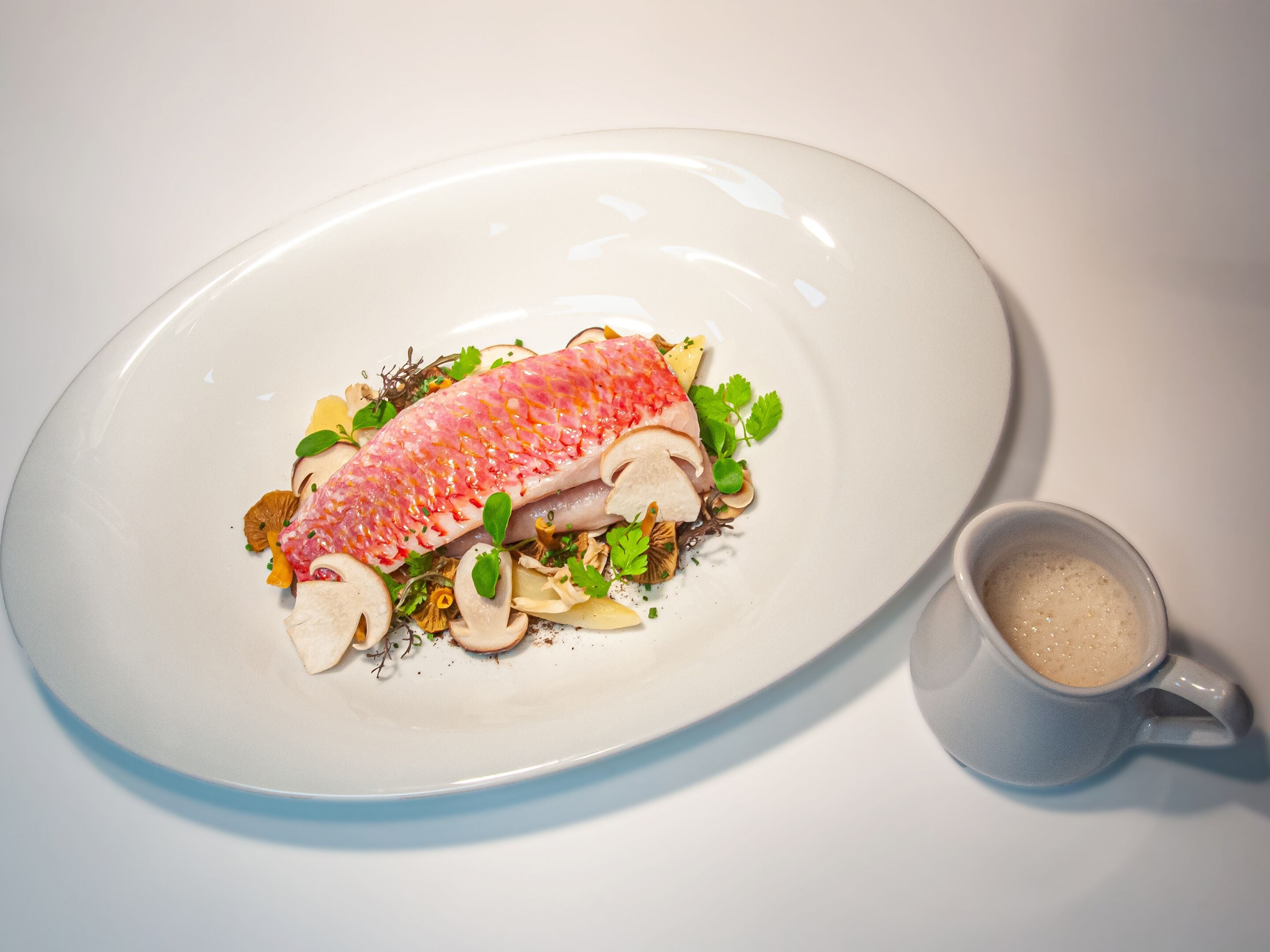 Um prato branco com peixe, cogumelos e ervas, e uma pequena chávena de café.