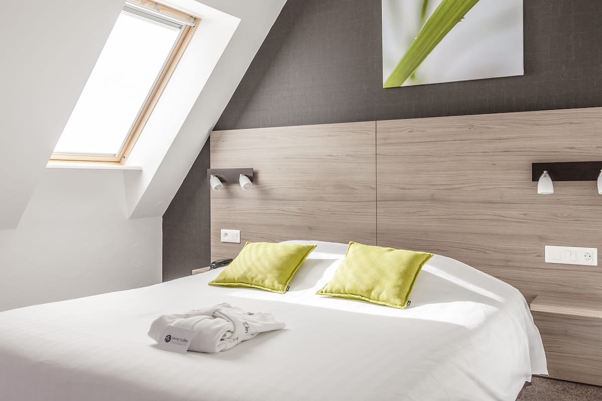 Quarto moderno com claraboia, cama com roupa de cama branca e almofadas verdes, cabeceira de madeira e luminária fixada na parede.