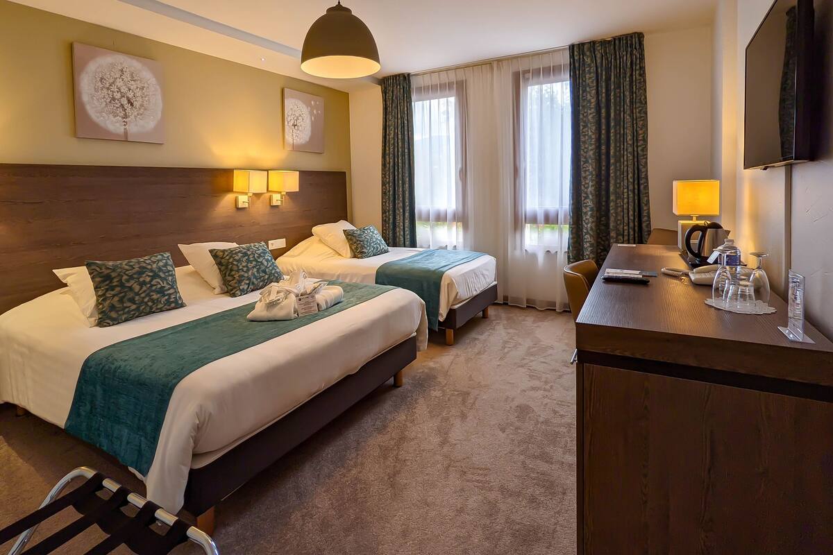 Quarto de hotel com duas camas, uma escrivaninha, uma cadeira, uma televisão e uma janela grande com cortinas.