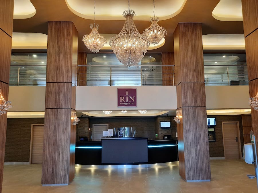 Área de recepção do RIN Airport Hotel com design moderno, apresentando um balcão central, grandes pilares de madeira e um candelabro.