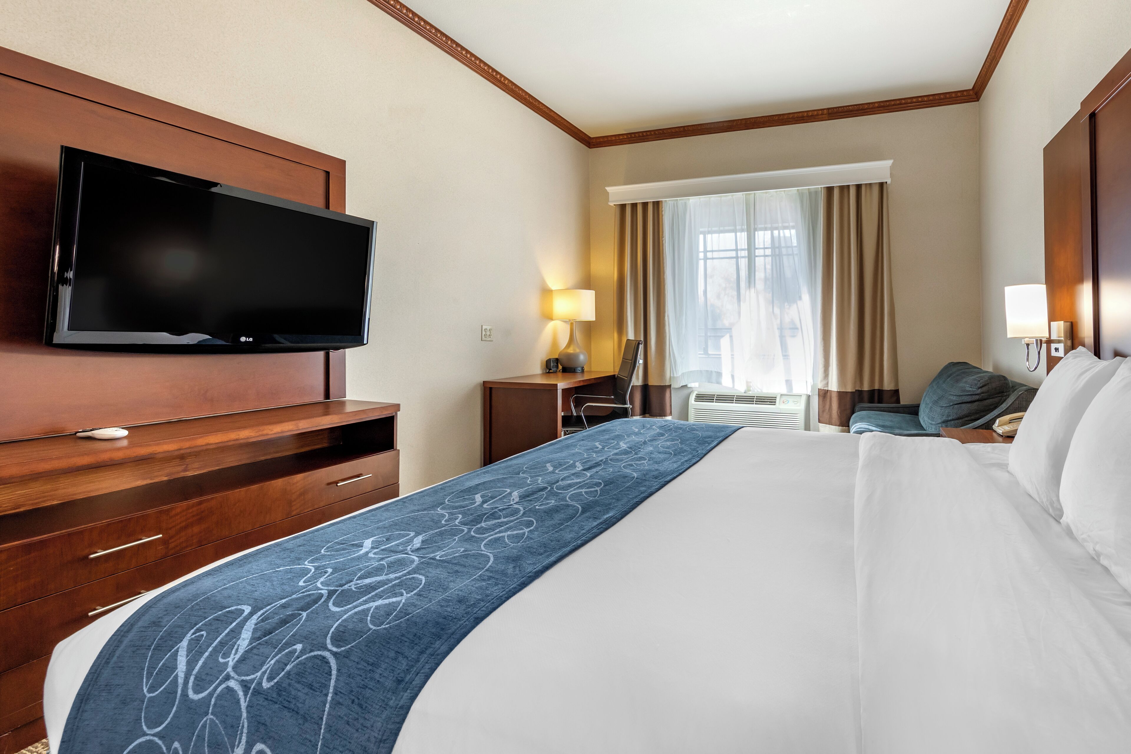 Quarto de hotel com uma cama grande, uma televisão de tela plana fixada em um armário de madeira, uma escrivaninha com luminária e uma janela com cortinas.